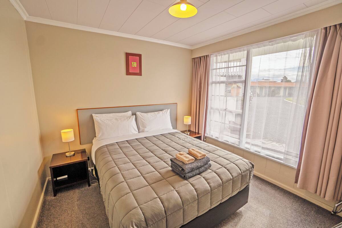 Quarto de hotel com uma cama grande, duas mesas de cabeceira com luminárias, uma janela com cortinas e um pequeno quadro na parede.