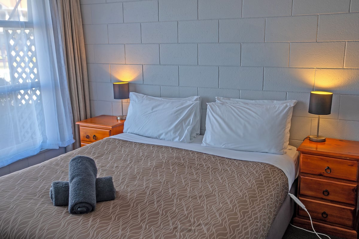 Quarto de hotel com uma cama grande, duas mesas de cabeceira com luminárias, um criado-mudo de madeira e uma janela com cortinas.