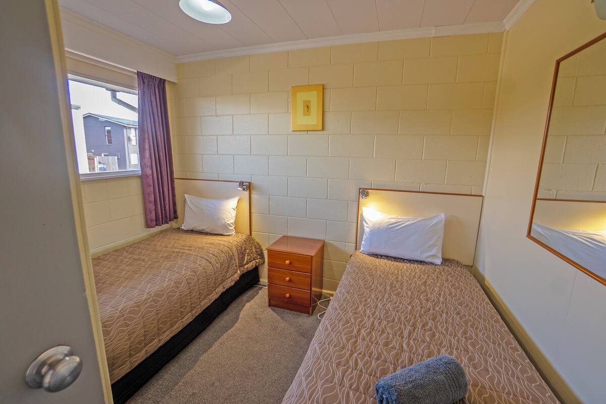Quarto com duas camas de solteiro, uma pequena mesa de cabeceira de madeira, janela com cortinas, quadro na parede e espelho no lado direito.
