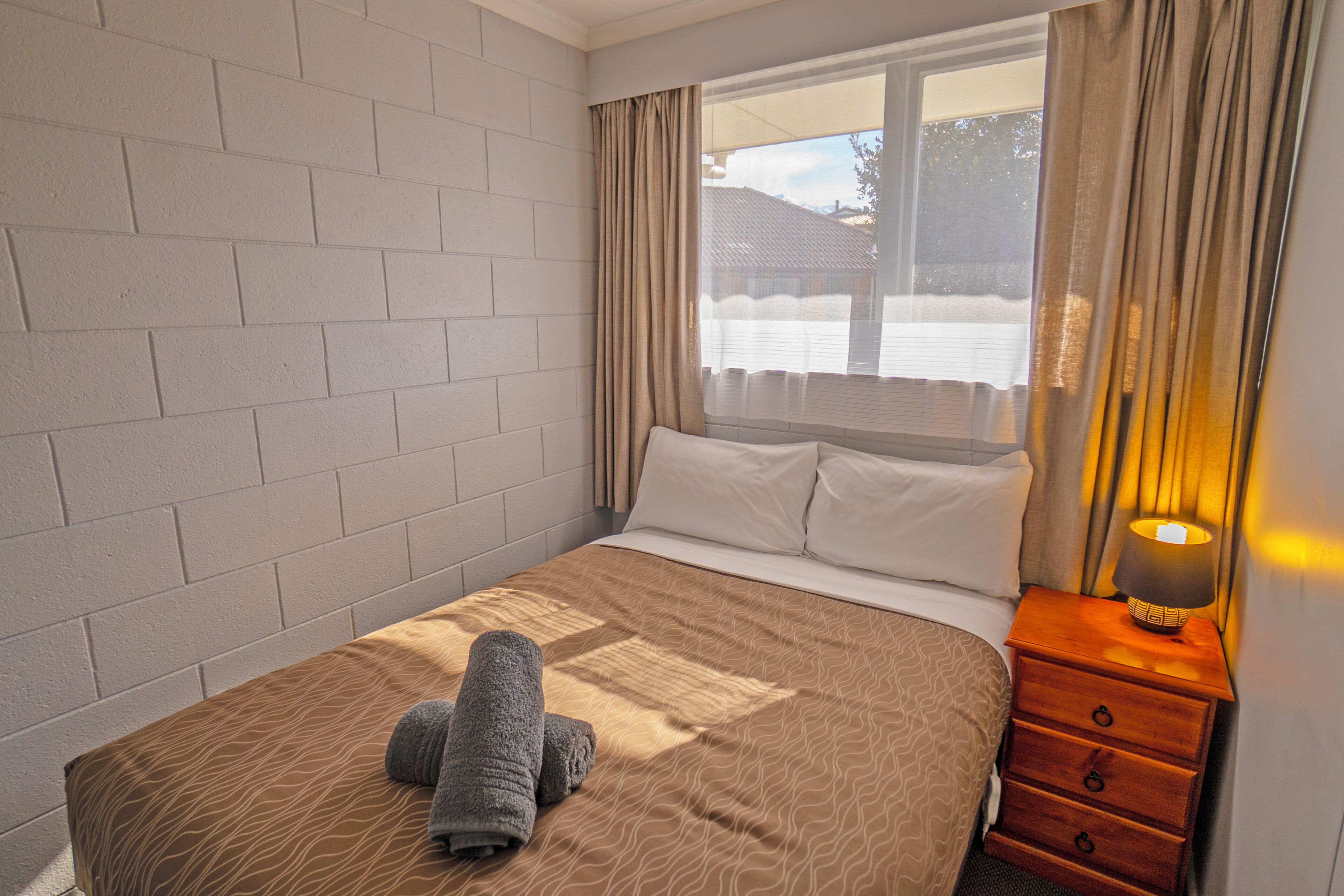 Quarto de hotel com cama, mesa de cabeceira e luminária, além de janela com cortinas.