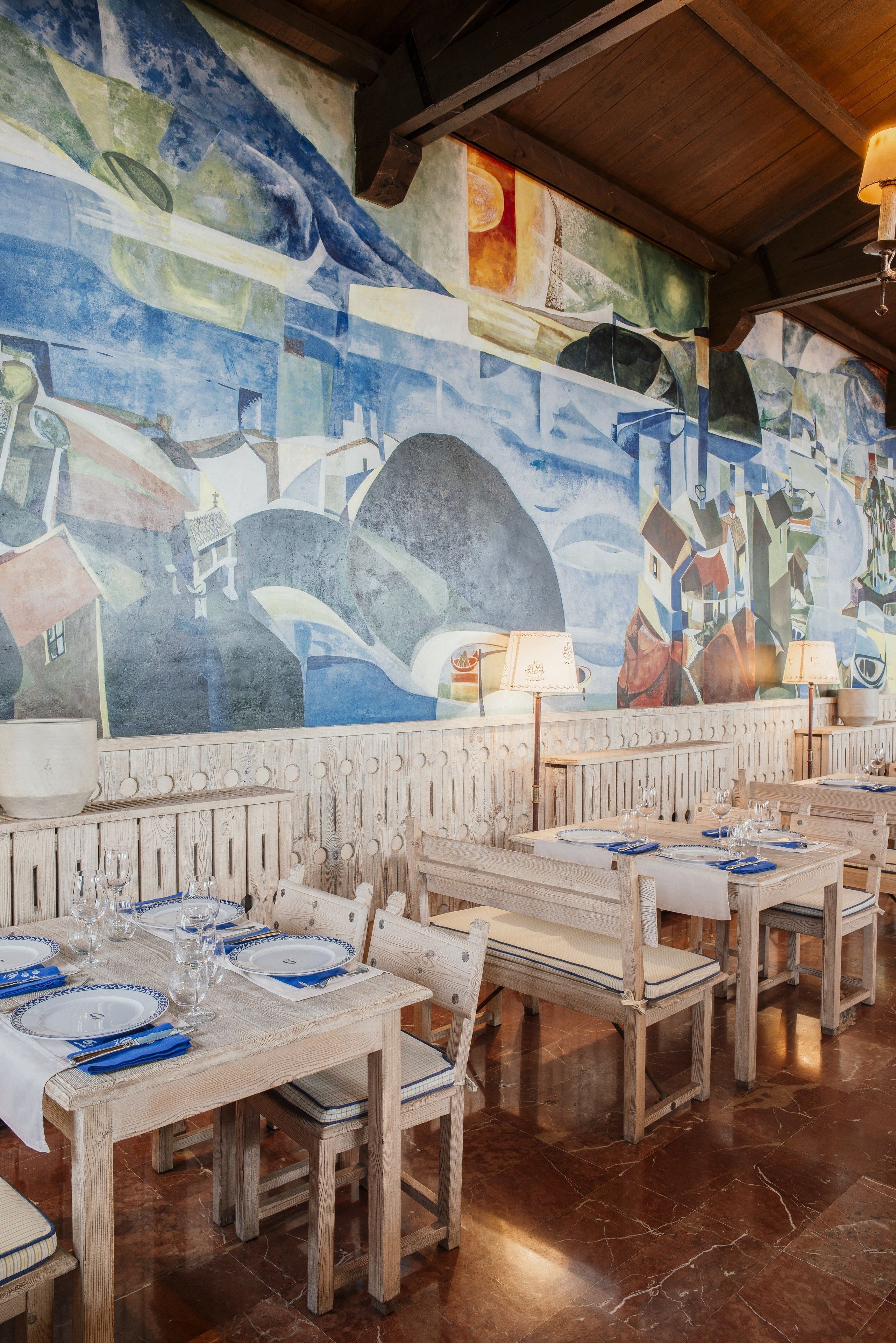 Um restaurante com mesas de madeira preparadas para refeições, um mural na parede e teto de madeira.