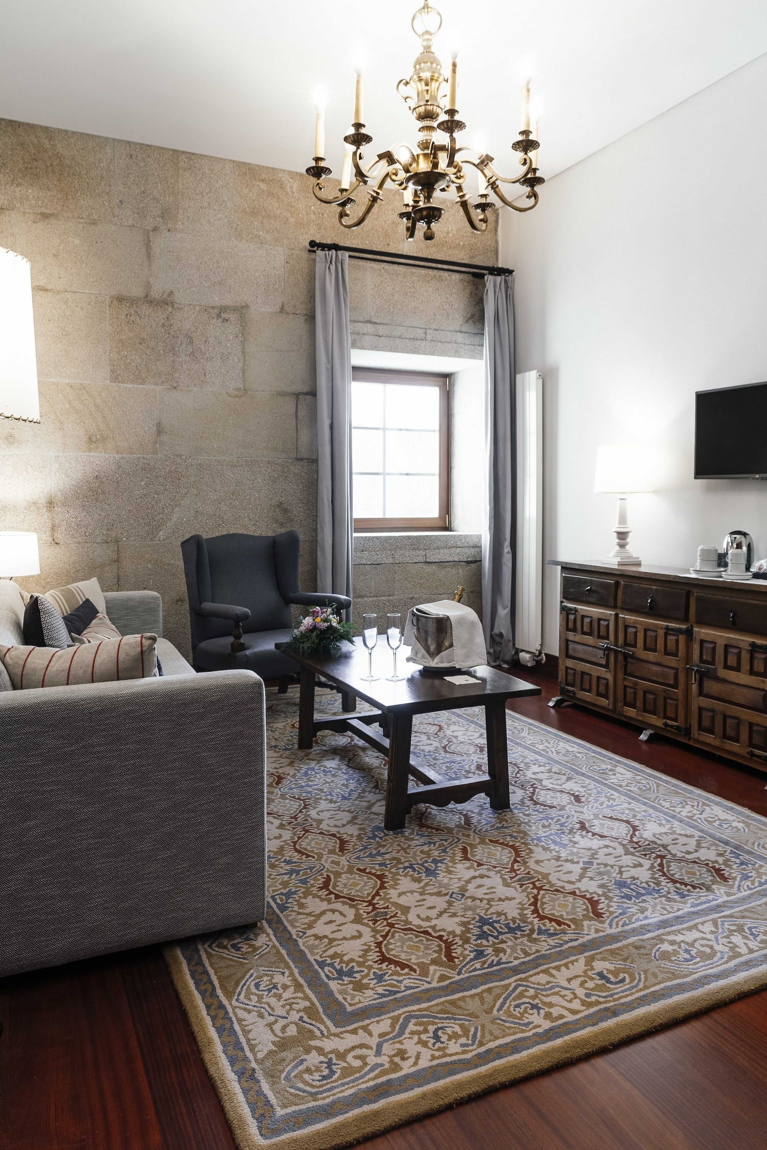 Uma sala de estar com parede de pedra, um candelabro, um sofá, uma poltrona, uma mesa de centro, uma TV e um armário.