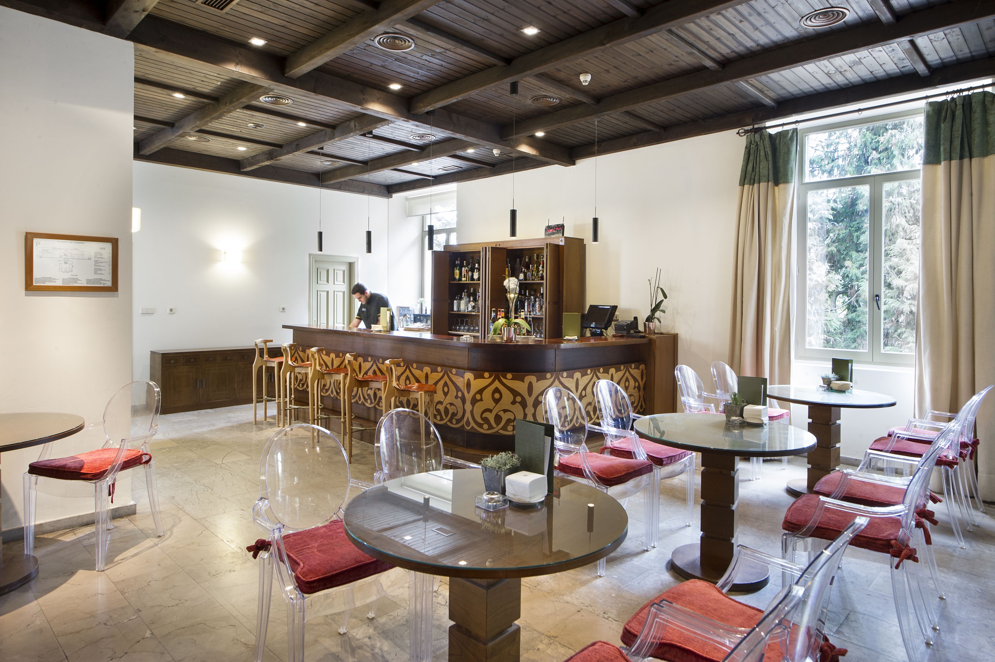 Restaurante com cadeiras transparentes, mesas redondas, um balcão de madeira e vista para árvores pela janela.
