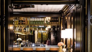 Uma área de bar com piso de mármore polido, banquetas, balcão com garrafas e um abajur decorativo.