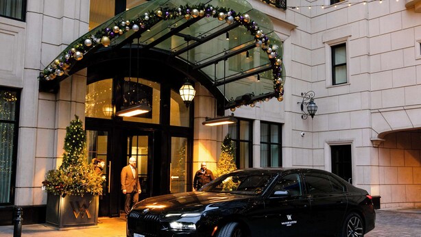 Um carro de luxo preto estacionado em frente à entrada de um hotel, com uma cobertura de vidro decorada com enfeites festivos.