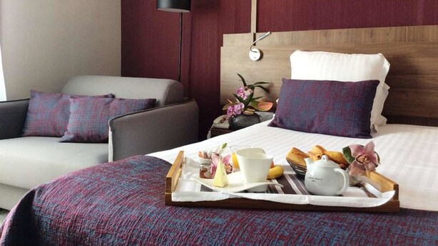 Quarto de hotel com cama, mesa de cabeceira com abajur, cadeira e bandeja com itens de café da manhã.