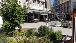 Um café ao ar livre com mesas e cadeiras, rodeado por vegetação e edifícios com varandas.