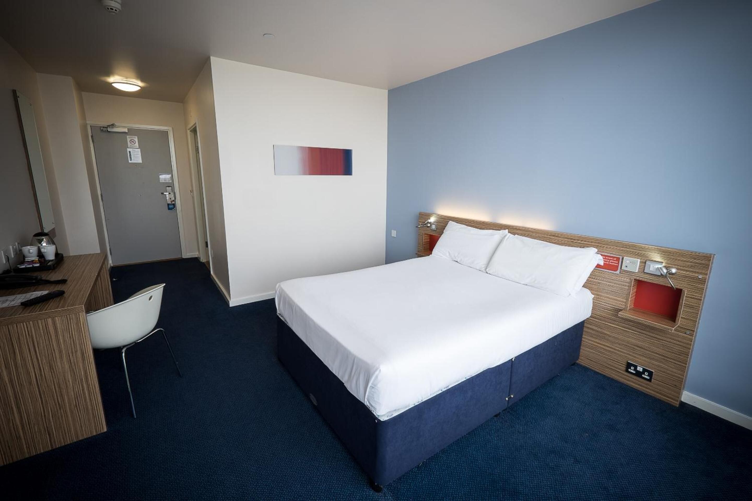 Quarto de hotel com cama, escrivaninha, cadeira e pequena mesa com prateleira vermelha.