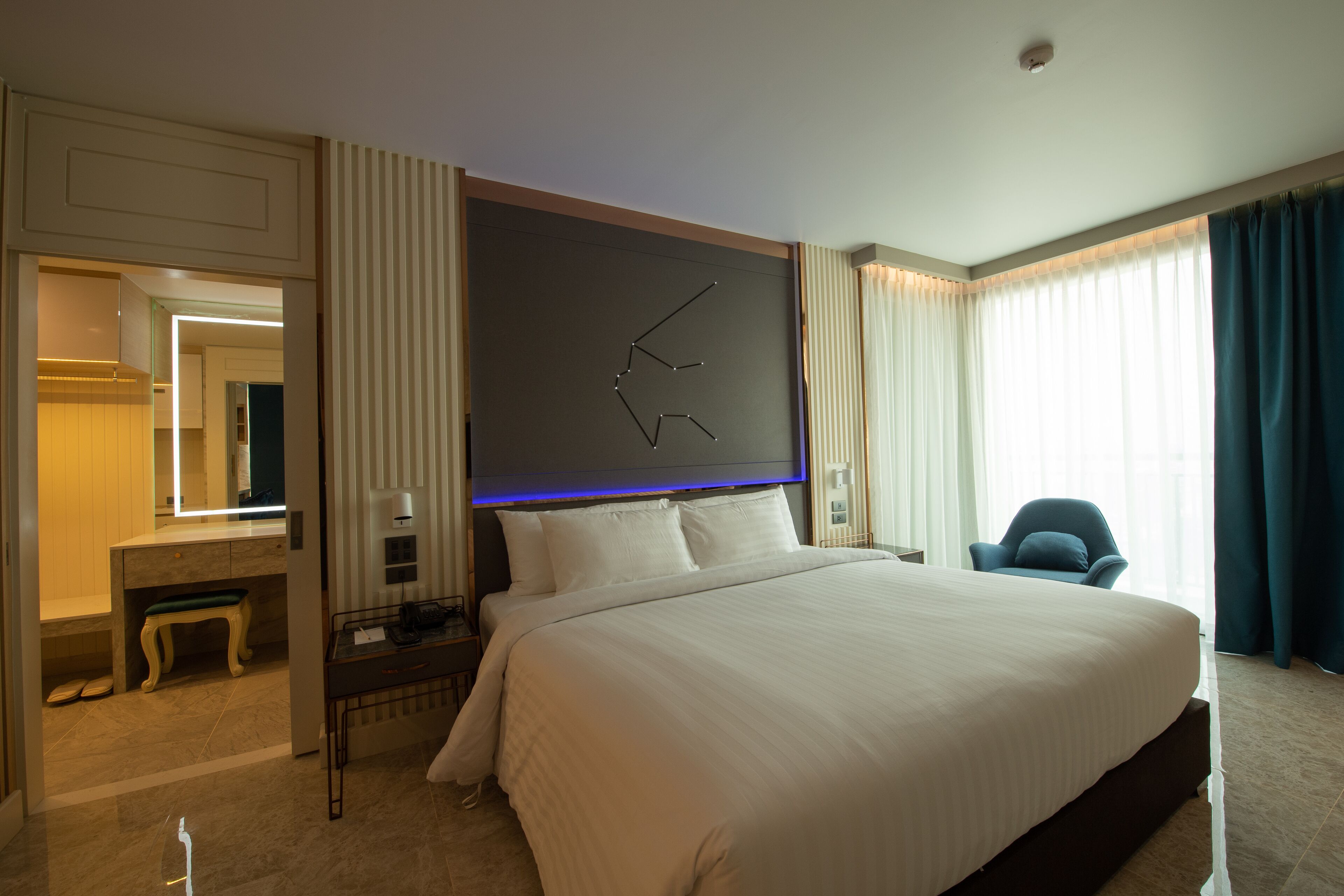 Quarto de hotel com uma cama grande, uma escrivaninha com cadeira, mesa de cabeceira, espelho e janela com cortinas.