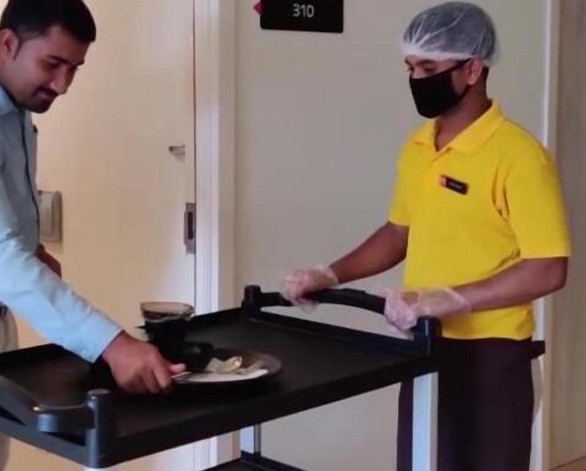 Quarto de hotel com uma mesinha preta e uma pessoa vestindo uma camisa amarela servindo comida.