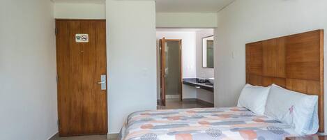 Quarto de hotel com uma cama, cabeceira de madeira, mesa de cabeceira, espelho e porta com placa de proibido fumar.