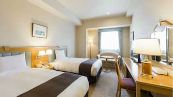 Quarto de hotel com duas camas, uma escrivaninha, uma cadeira, um abajur e uma janela com cortinas.