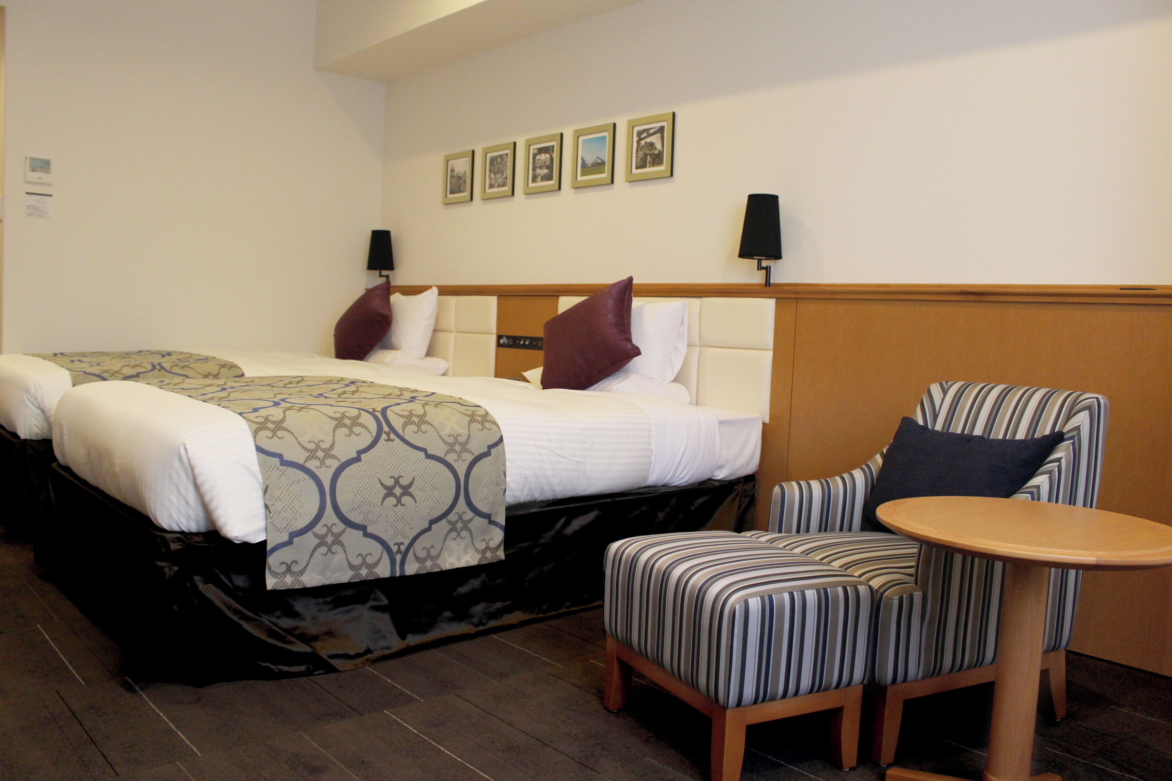 Quarto de hotel com duas camas, uma poltrona listrada, uma mesinha redonda e uma parede com quadros.