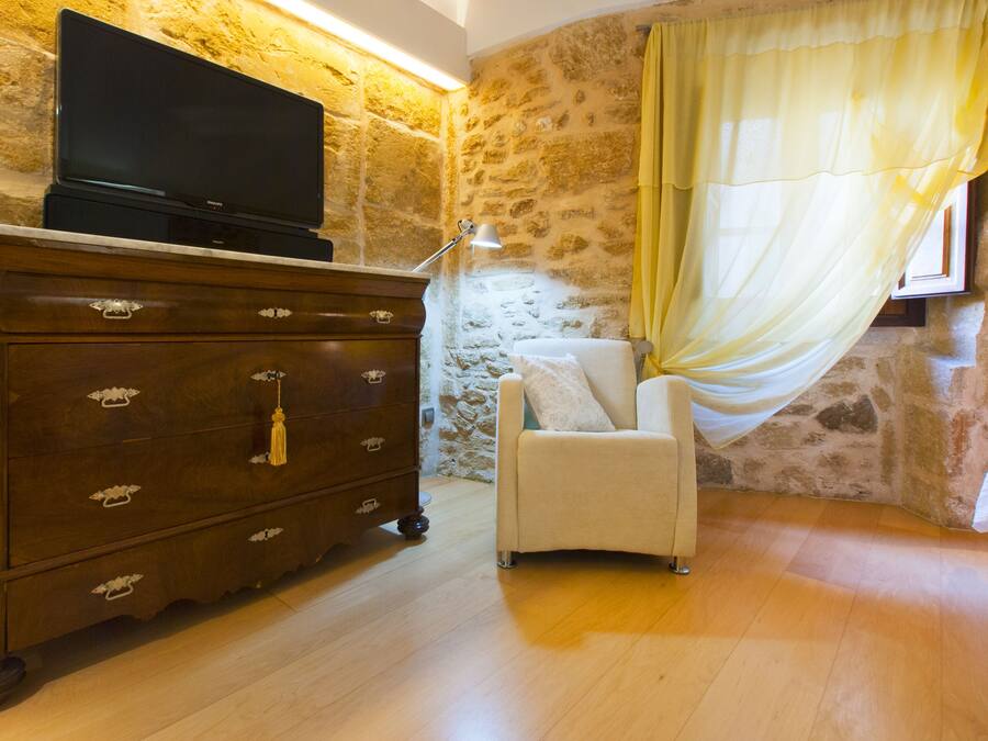 Quarto com parede de pedra, cômoda de madeira com gavetas, uma televisão em cima, uma poltrona branca e uma janela com cortinas amarelas.