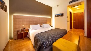 Quarto de hotel com uma cama grande, uma mesinha de cabeceira, um banco e uma porta que dá para outro quarto.