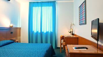 Quarto de hotel com cama, escrivaninha, cadeira, televisão e janela com cortinas.