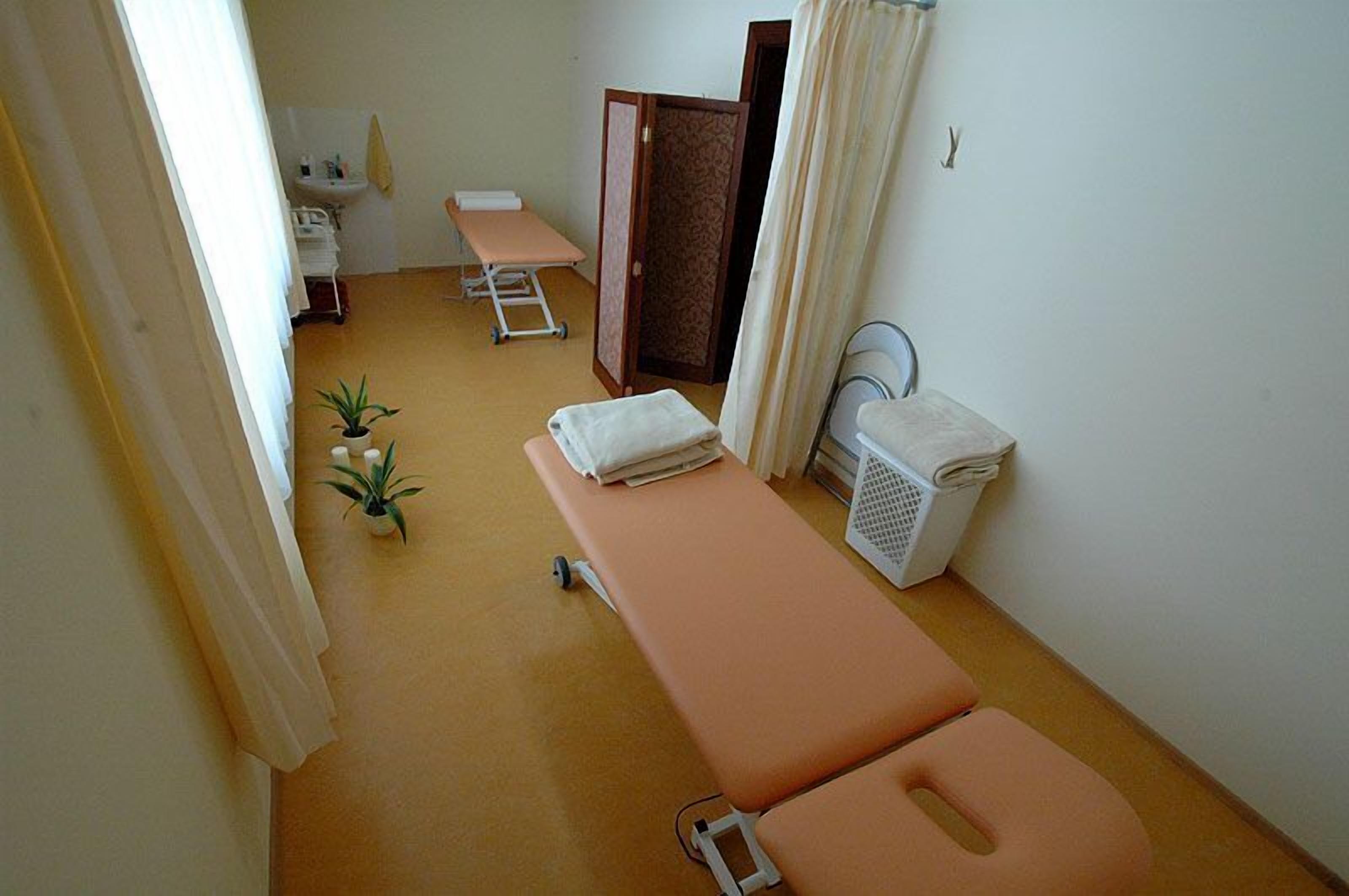 Uma sala de exames médicos com uma maca, uma pia pequena e uma janela com cortinas.