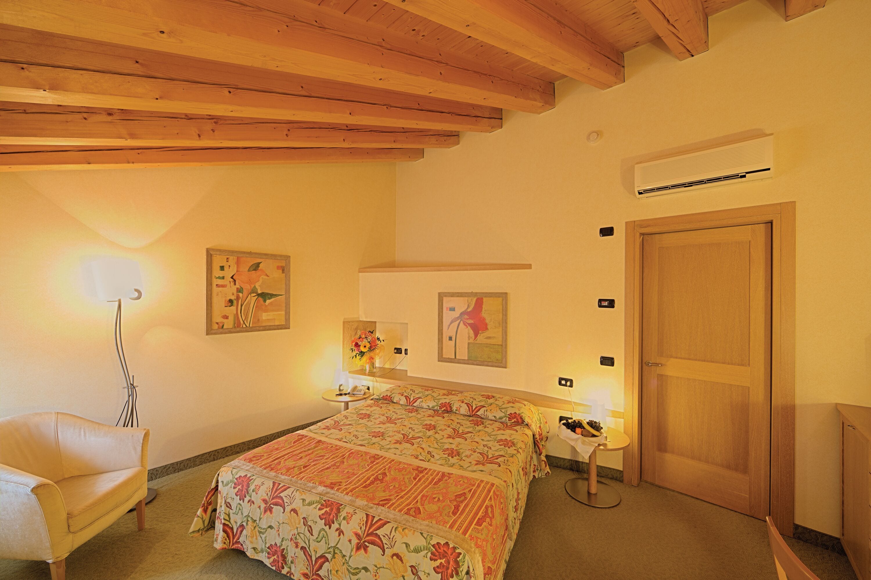Quarto de hotel com cama, uma cadeira, escrivaninha com luminária e porta.