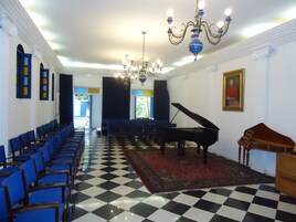 Um salão imponente com piso xadrez, fileiras de cadeiras azuis, um piano de cauda e um candelabro.