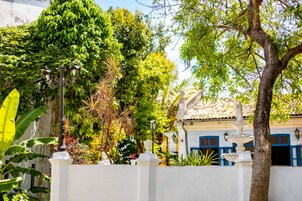 Um edifício branco com persianas azuis, rodeado por vegetação exuberante e uma parede branca com uma fonte decorativa.