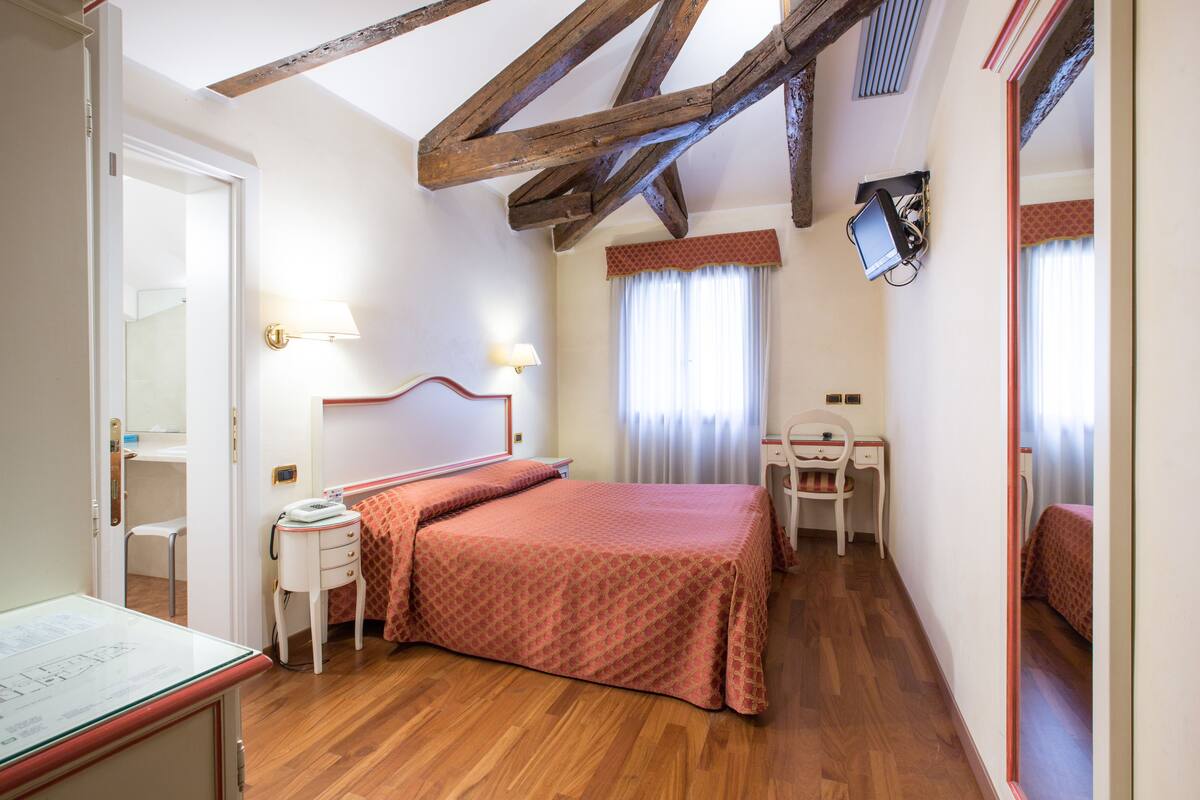Quarto com teto de madeira, cama com colcha estampada, escrivaninha e uma cadeira.