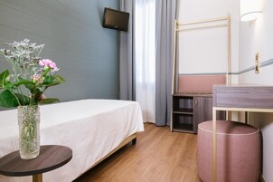 Quarto de hotel com cama, uma mesinha com um vaso de flores, televisão na parede e escrivaninha com cadeira.