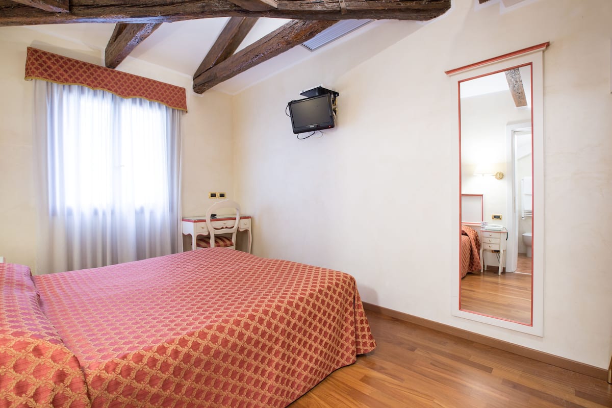 Quarto com cama, escrivaninha, cadeira, televisão e espelho.