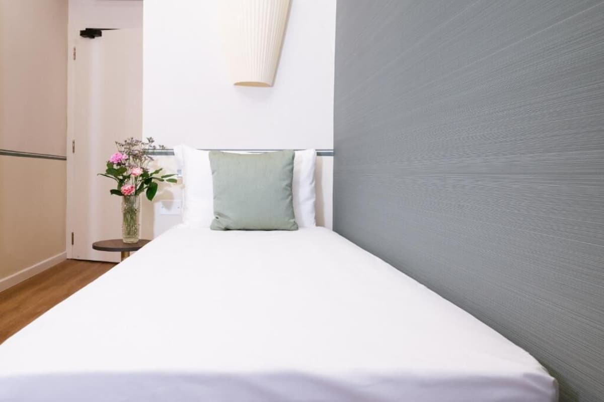 Uma cama de solteiro com colchão branco e travesseiro verde, um vaso com flores e uma luminária fixada na parede.