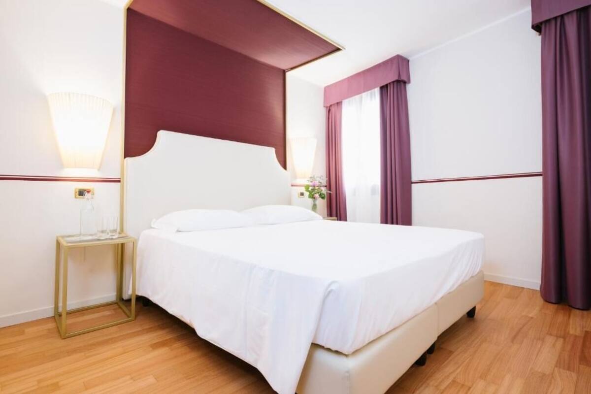 Uma cama bem arrumada em quarto com piso de madeira, mesa de cabeceira com abajur e uma janela grande com cortinas.