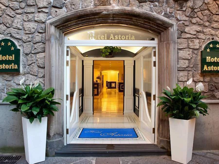 Entrada do Hotel Astoria, com fachada de pedra, um tapete azul na entrada e vasos de plantas em ambos os lados.