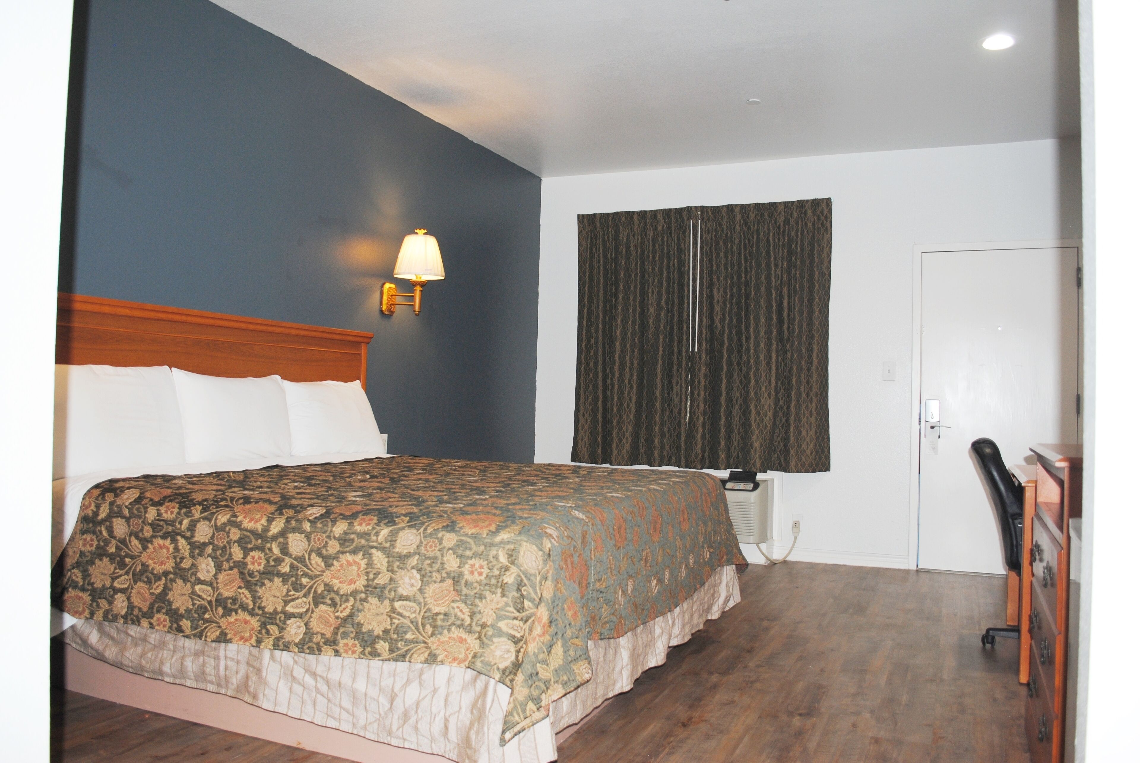 Um quarto com cama, mesa de cabeceira, abajur, janela com cortinas e porta.