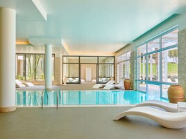 Piscina interior moderna com paredes de vidro transparente, cadeiras de lounge brancas e vista para o exterior.