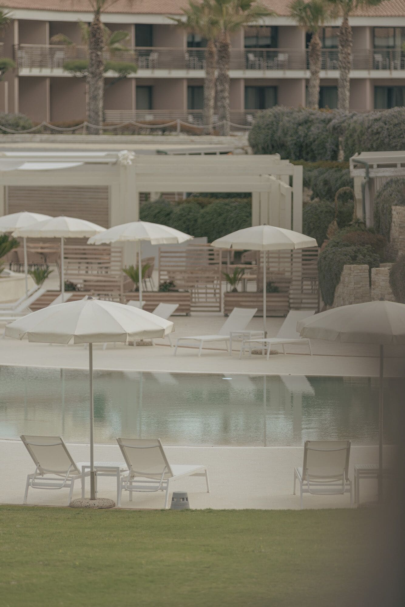 Área de piscina com espreguiçadeiras e guarda-sóis, cercada por um edifício com varandas e palmeiras.