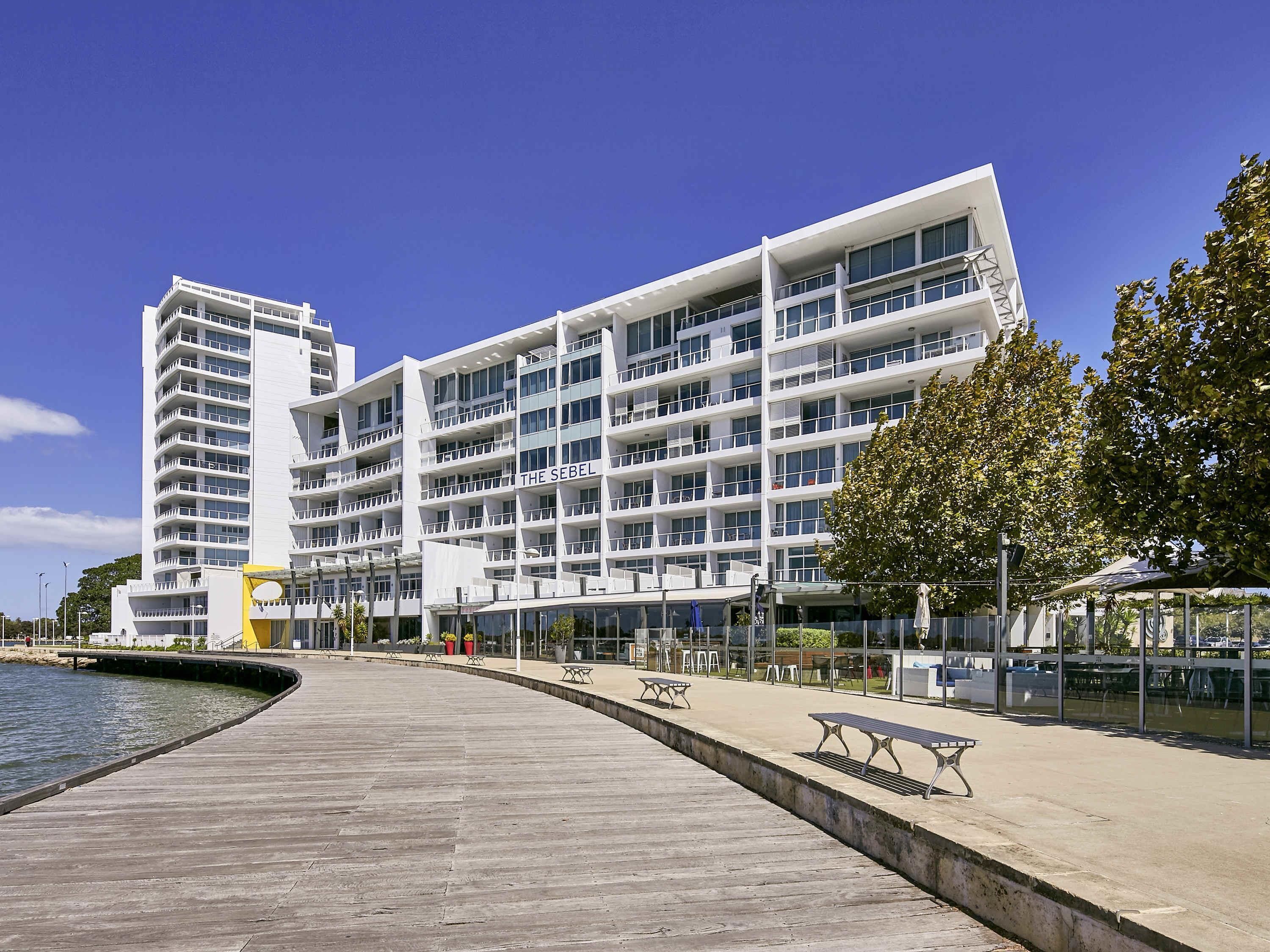 Um hotel moderno de vários andares, com varandas, calçadão à beira-mar e áreas de estar ao ar livre.