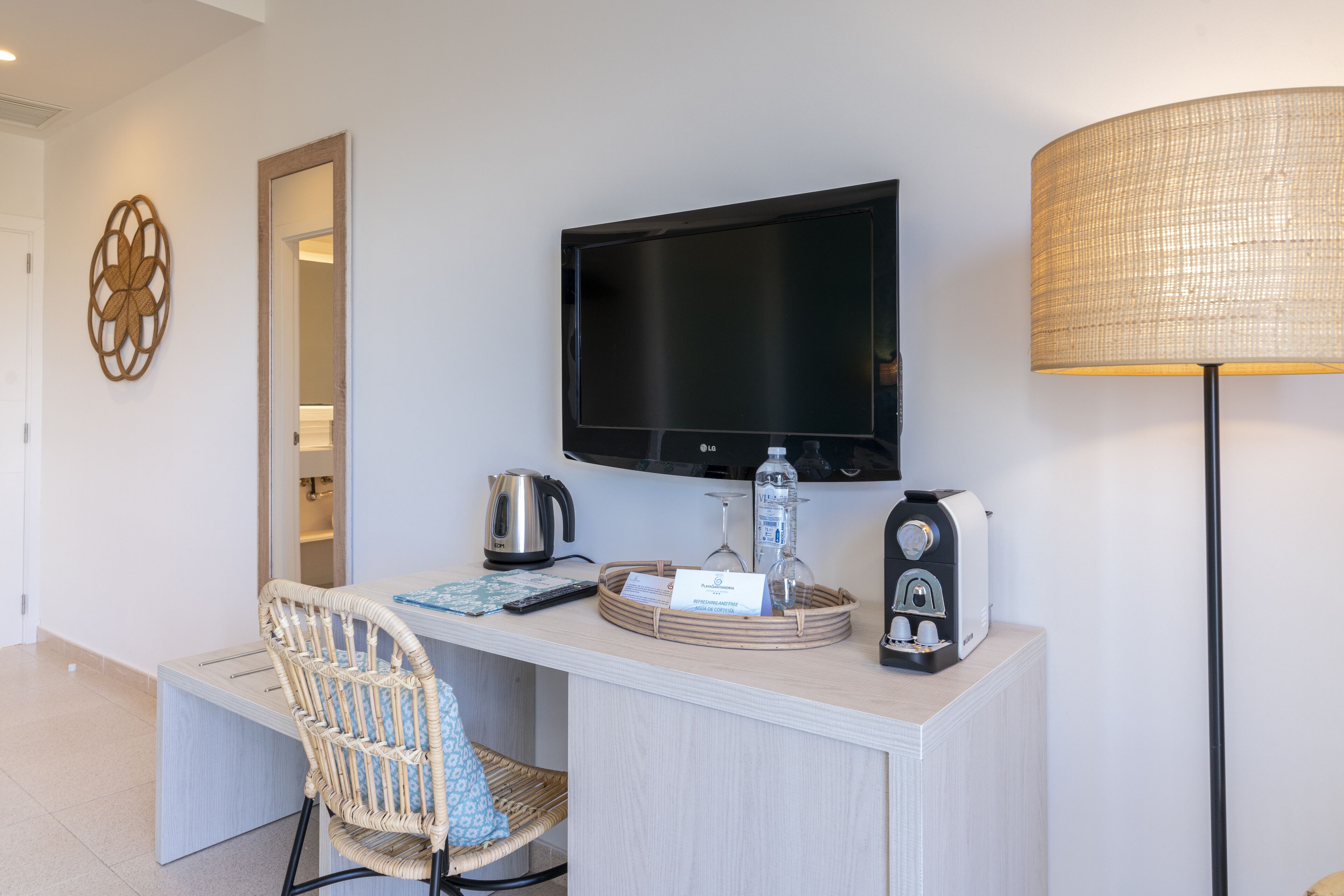 Quarto de hotel moderno com televisão de tela plana, máquina de café, chaleira, cadeira de vime e mesa de cabeceira com abajur.
