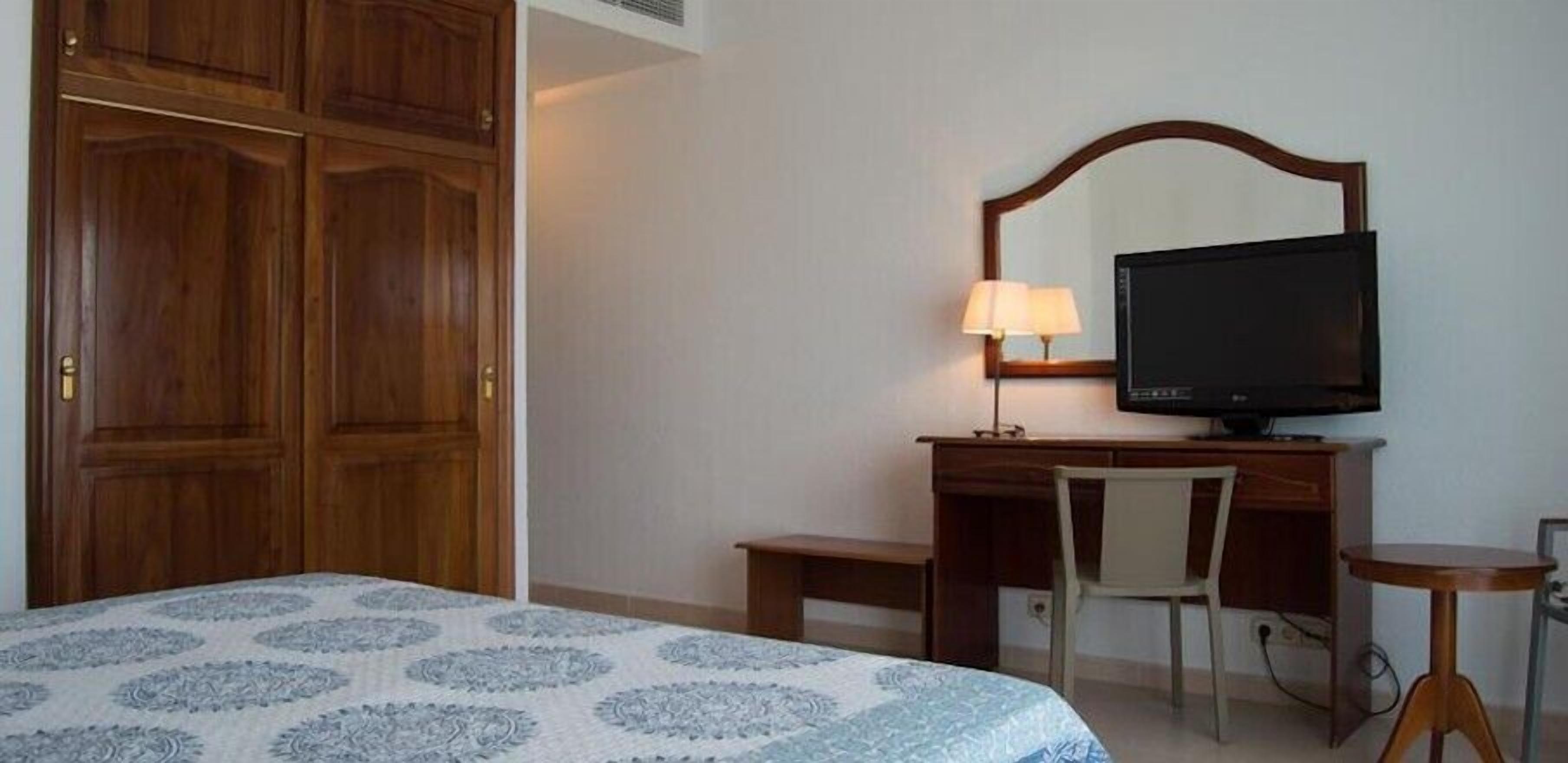 Quarto de hotel com uma cama, uma secretária, uma cadeira, uma televisão e um guarda-roupa.