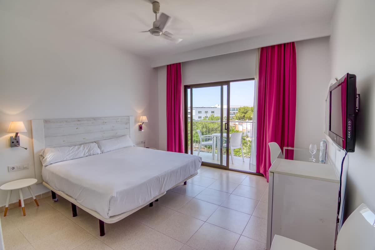 Quarto de hotel com cama, escrivaninha, televisão e varanda com cortinas cor-de-rosa.