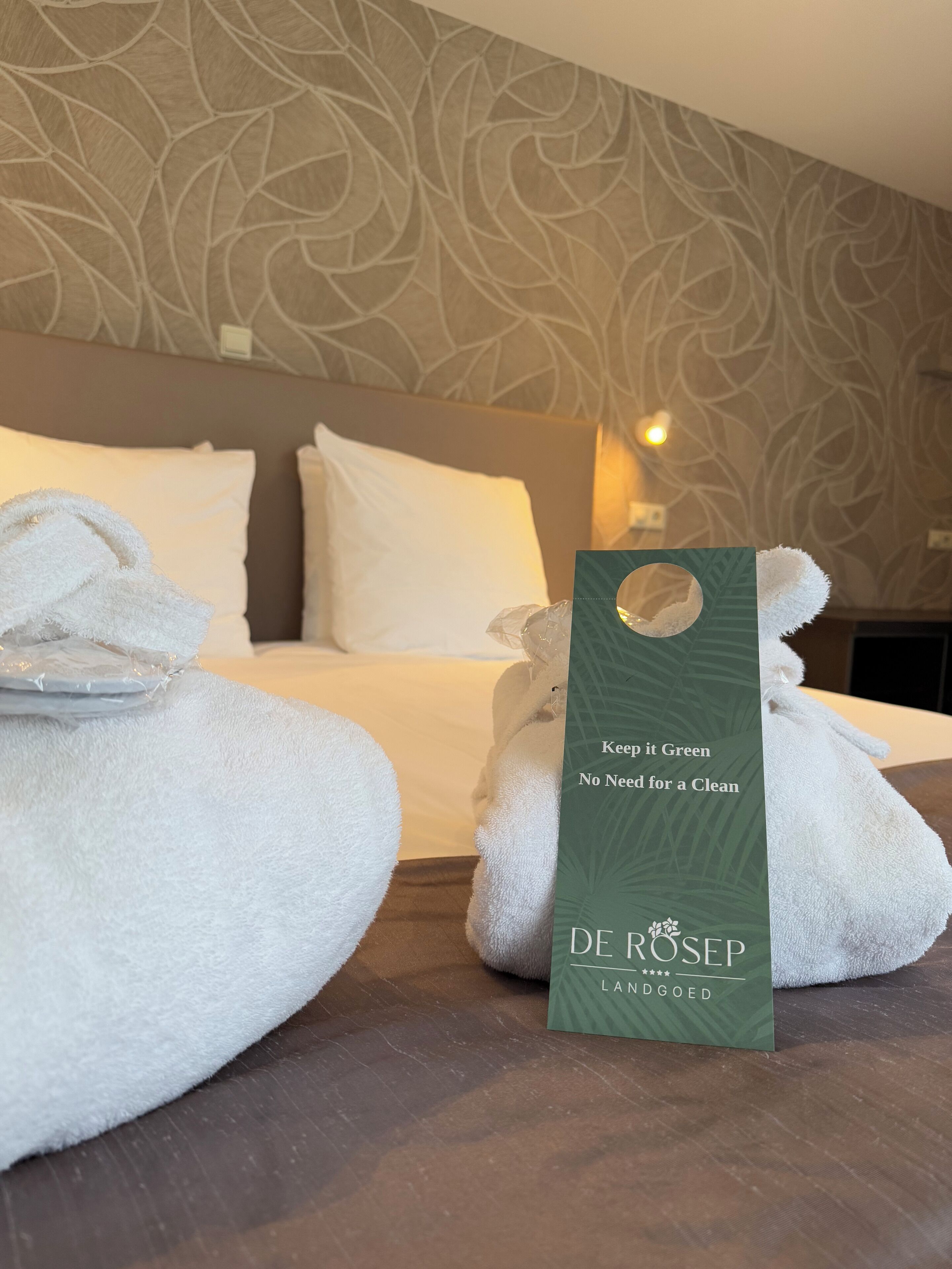 Quarto de hotel com cama bem arrumada, duas toalhas brancas e um sinal verde no chão.