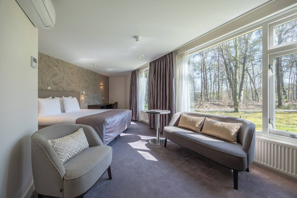 Quarto de hotel com cama, sofá, poltrona, mesa pequena e janelas amplas com vista para árvores.