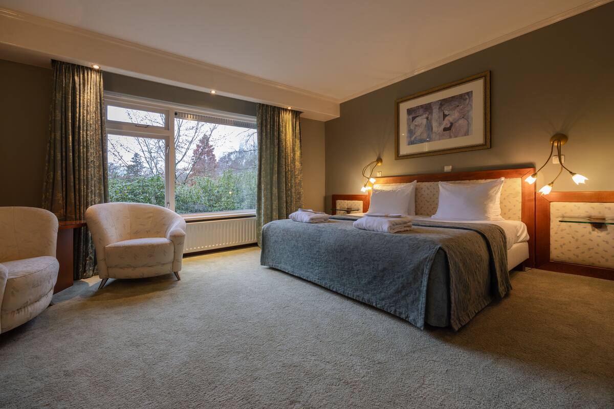 Quarto de hotel com uma cama grande, duas poltronas, uma janela com cortinas, um quadro na parede e uma mesa de cabeceira com abajur.