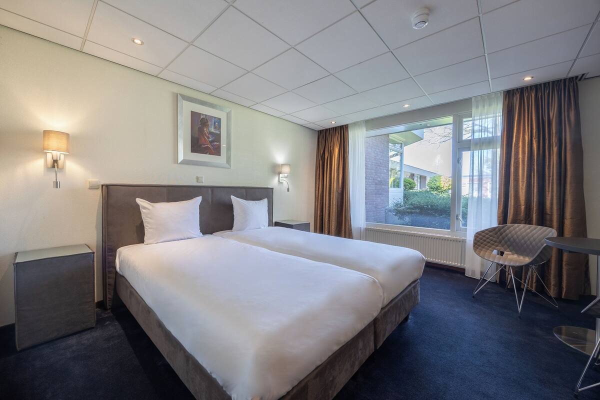 Quarto de hotel com cama de casal, duas mesas de cabeceira, uma cadeira, uma mesinha e uma janela com cortinas.