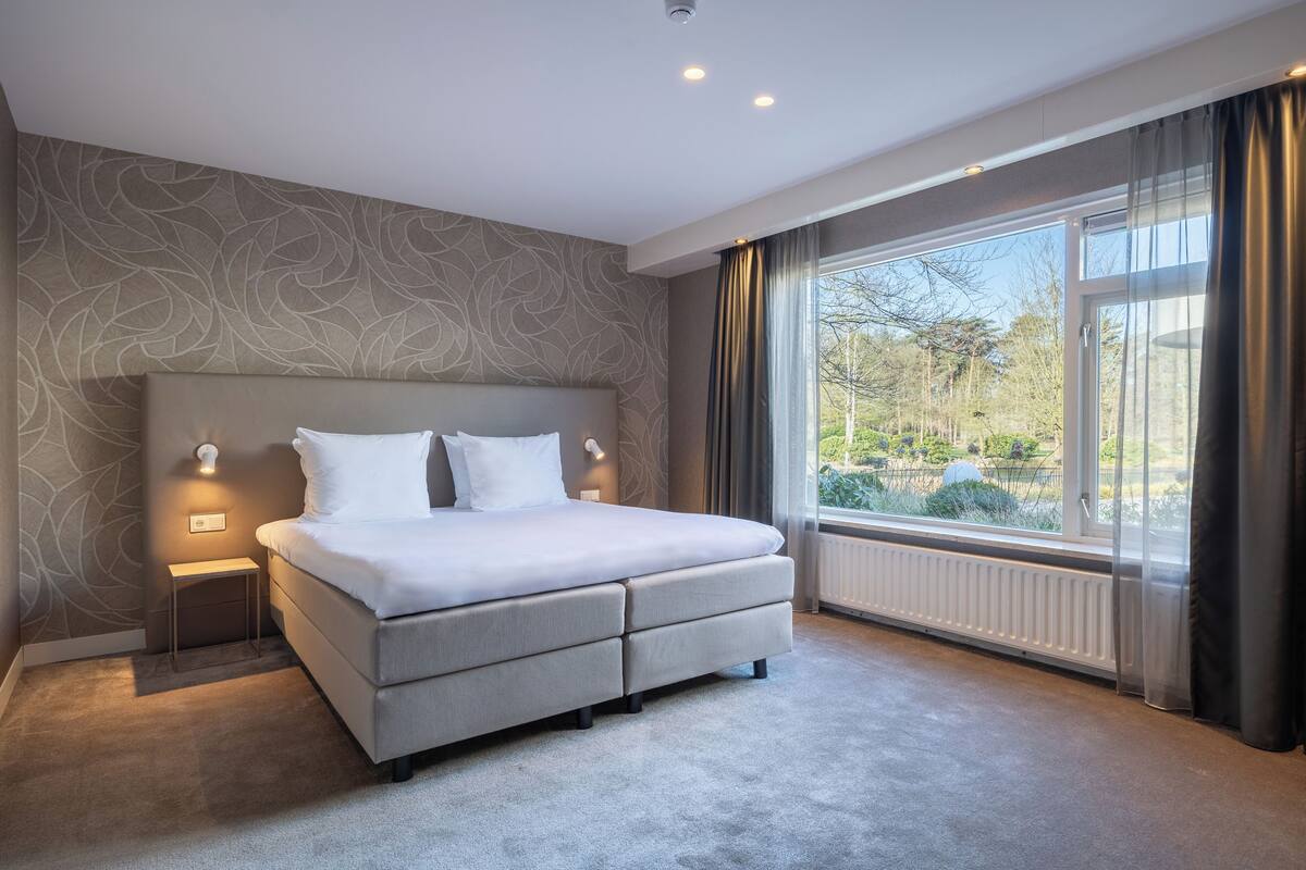 Quarto moderno com cama grande, mesa de cabeceira, janela com cortinas e vista para o verde lá fora.