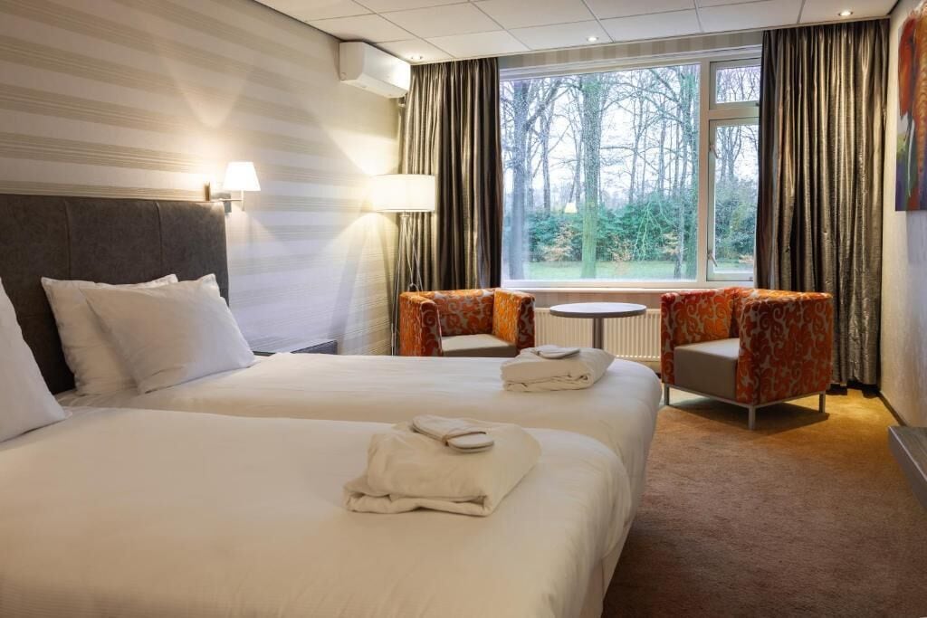 Quarto de hotel com duas camas, uma mesinha, duas cadeiras e janela com vista para árvores.