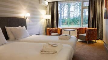 Quarto de hotel com duas camas, uma mesinha, duas cadeiras e janela com vista para árvores.