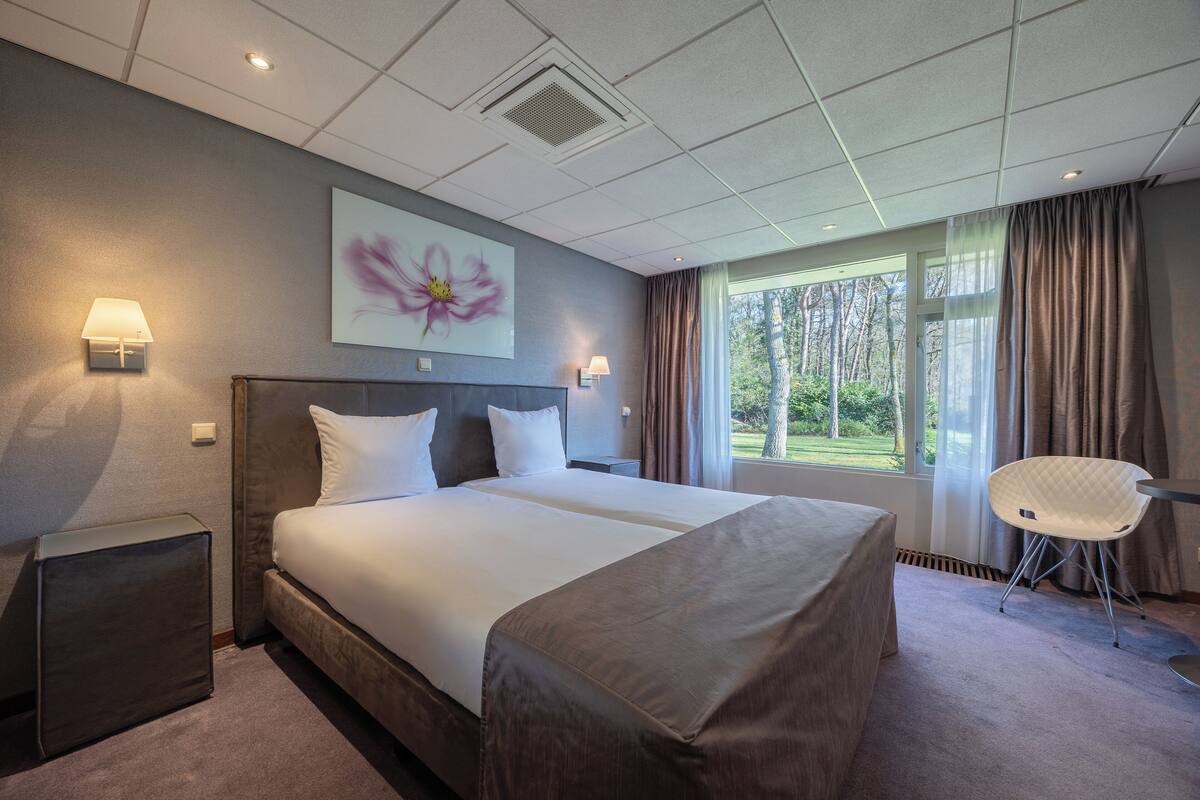 Quarto de hotel com uma cama grande, duas mesas de cabeceira, uma cadeira, uma janela com cortinas e um quadro de flores na parede.