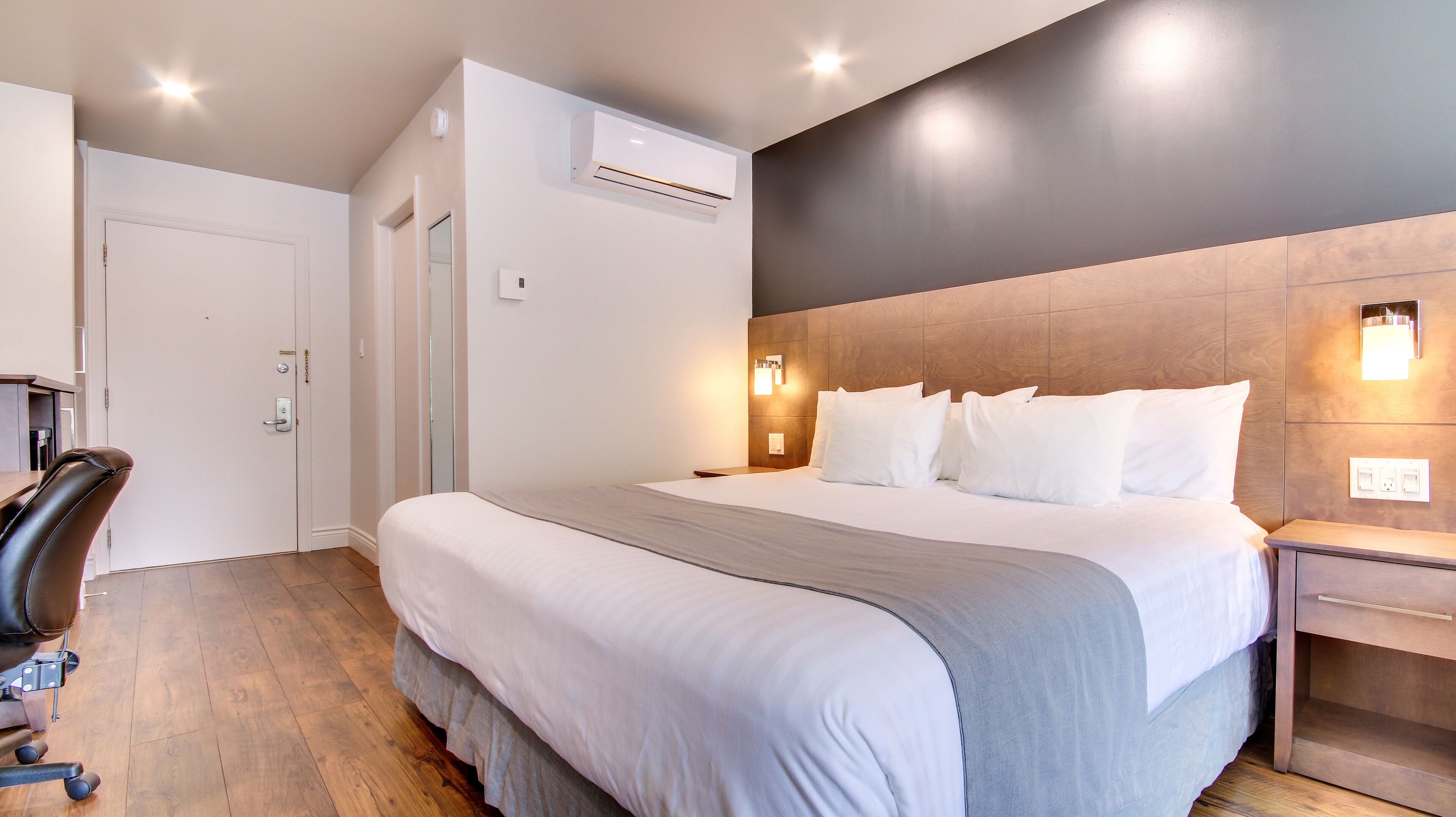 Quarto de hotel com uma cama grande, uma escrivaninha com cadeira, um aparelho de ar condicionado e uma mesa de cabeceira com abajur.