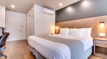 Quarto de hotel com uma cama grande, uma escrivaninha com cadeira, um aparelho de ar condicionado e uma mesa de cabeceira com abajur.