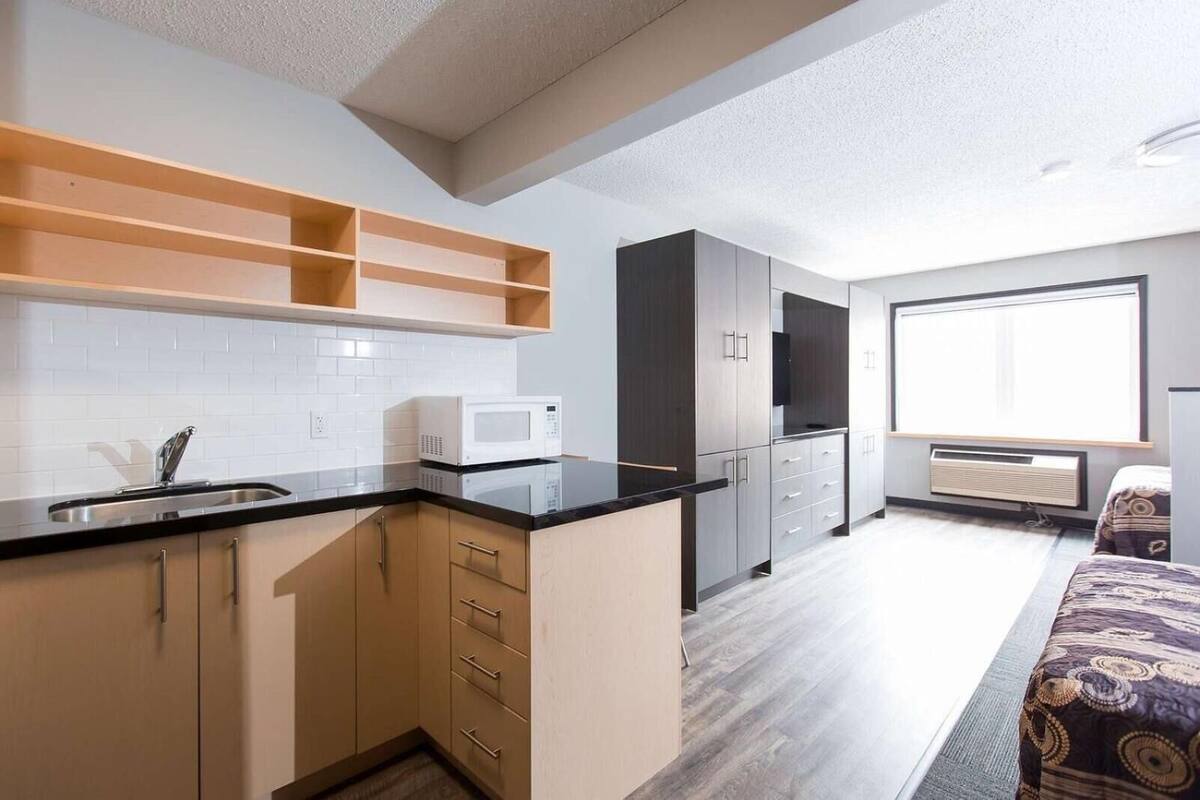 Uma cozinha moderna com micro-ondas, pia e armários de madeira. Há uma janela que traz luz natural e uma cama com roupa de cama estampada no quarto adjacente.