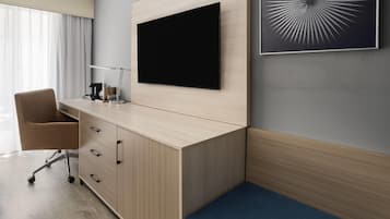 Quarto de hotel moderno com uma grande TV de tela plana na parede, uma escrivaninha com cadeira, uma pequena mesa de cabeceira e um banco estofado azul.