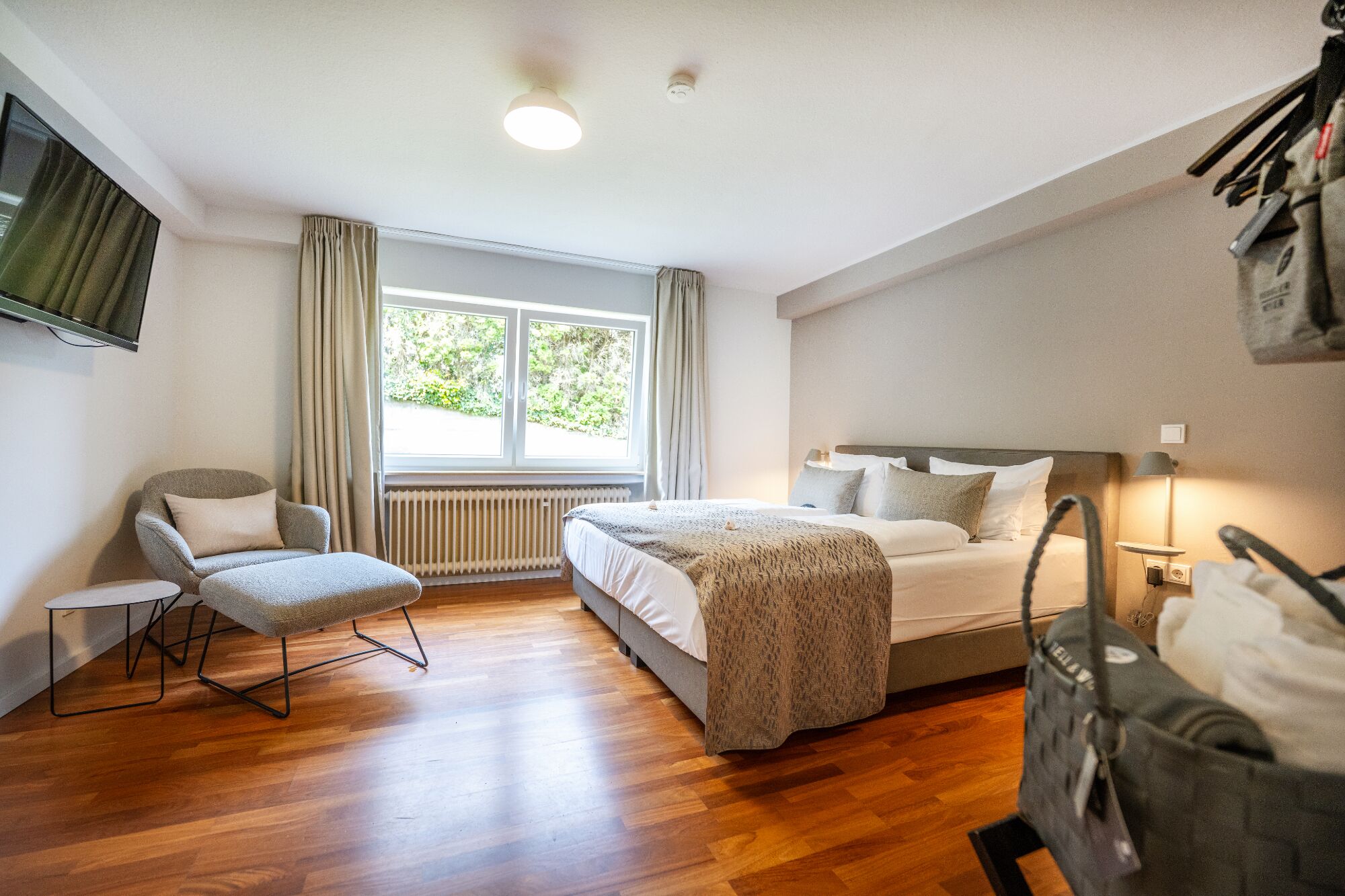 Quarto de hotel com cama, poltrona, janela, radiador e televisão fixada na parede.